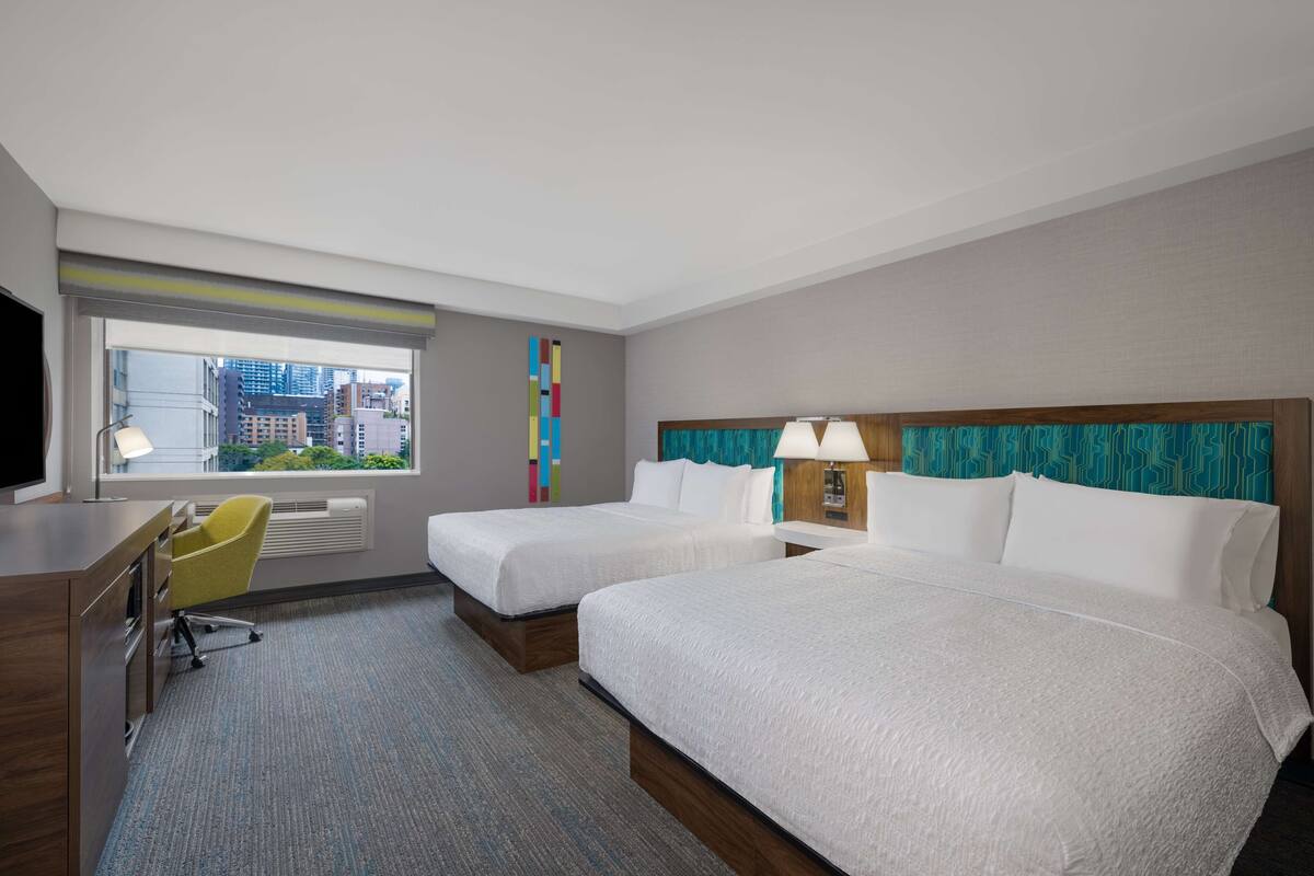Quarto de hotel com duas camas, uma escrivaninha, uma cadeira e janela com vista para a cidade.
