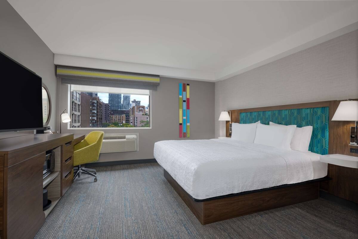 Quarto de hotel com uma cama grande, uma escrivaninha com cadeira, televisão e janela com vista para a cidade.