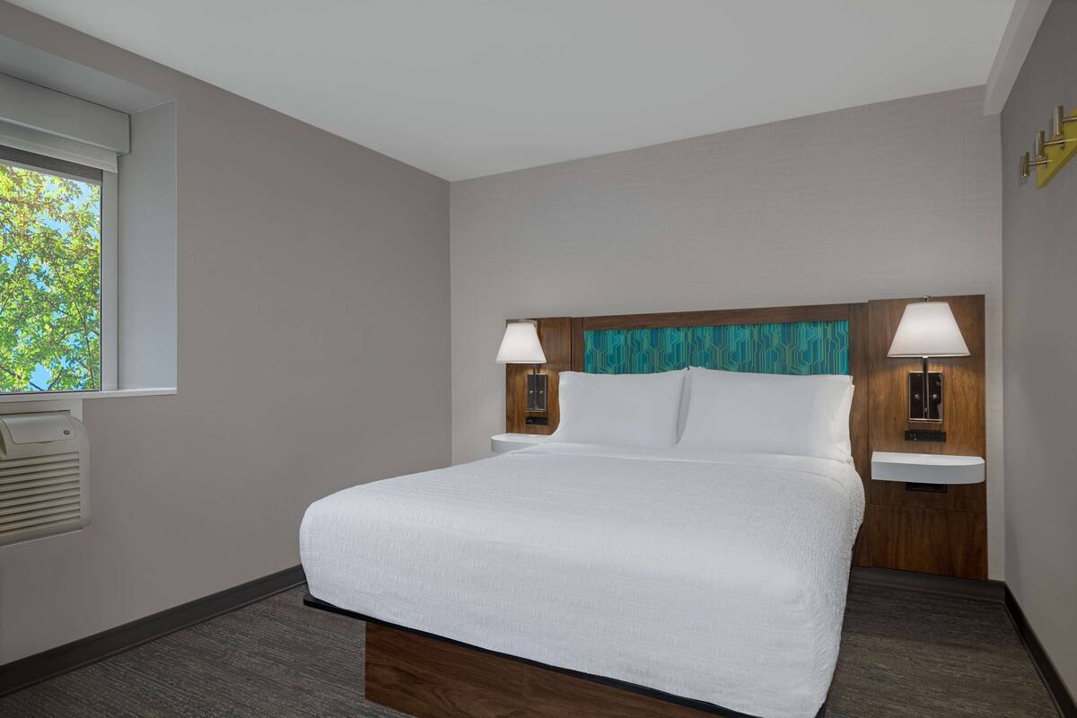 Quarto de hotel com uma cama grande, duas mesas de cabeceira, uma janela com persianas, um ar condicionado fixado na parede e um abajur na parede.