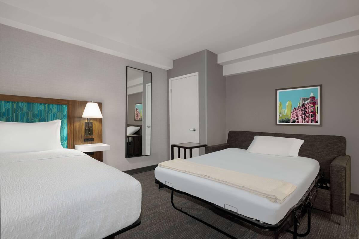 Quarto de hotel com duas camas, uma escrivaninha, uma cadeira, um abajur, um espelho e uma pintura da paisagem urbana.