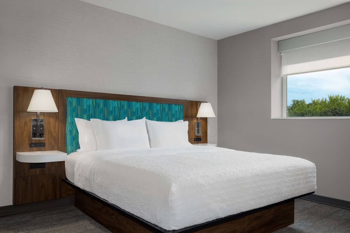 Quarto de hotel com uma cama grande, duas luminárias de cabeceira, um painel de cabeceira de madeira e uma janela com vista para o verde.