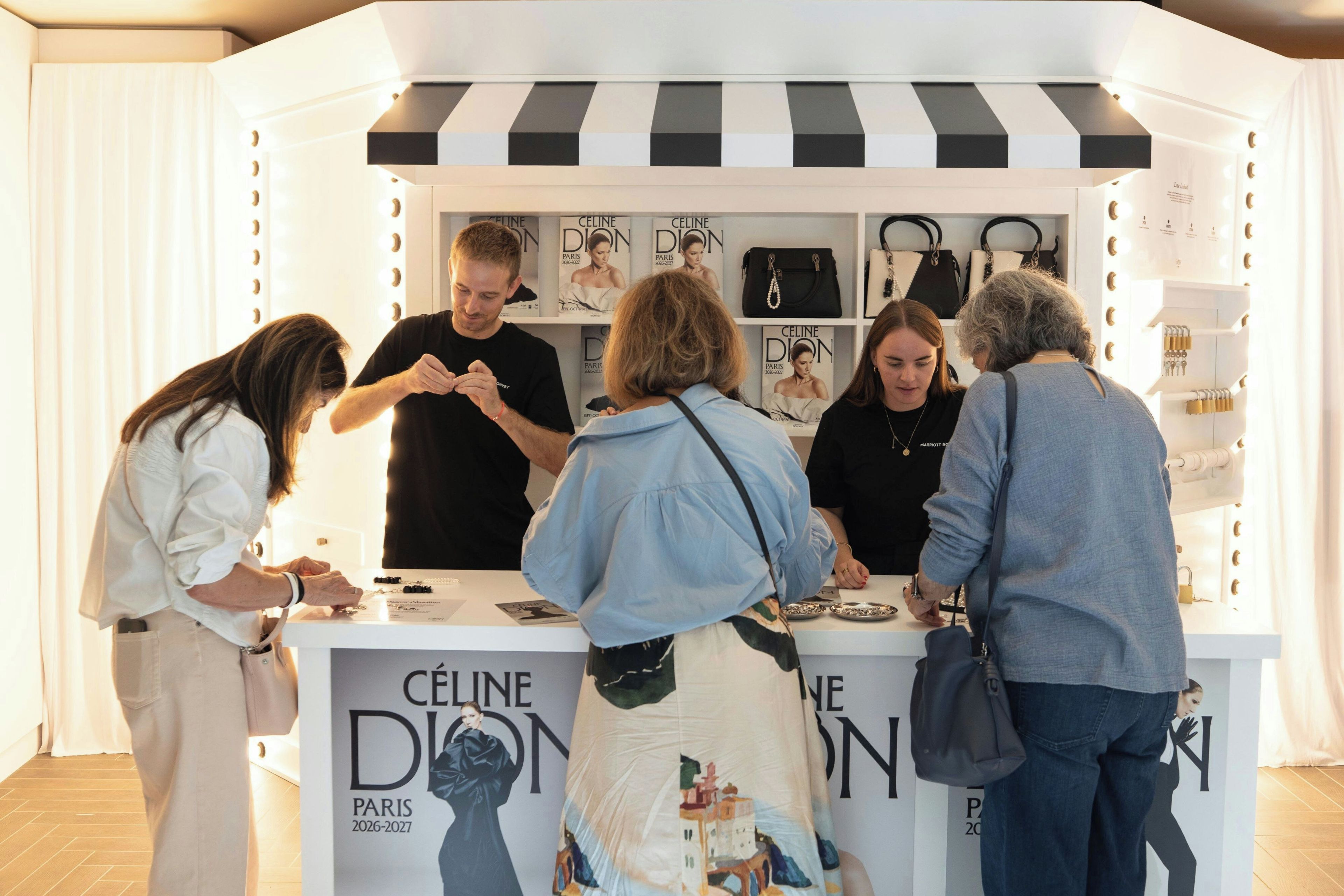 Um grupo de pessoas em um evento da Celine Dion Paris 2026-2027, interagindo com uma exposição de bolsas e acessórios.