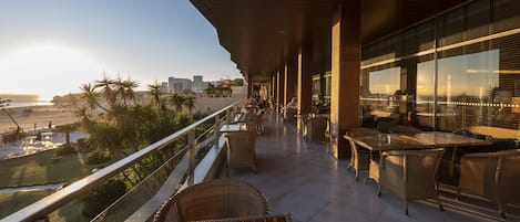 Varanda com mobiliário de vime, vista para a praia e paisagem urbana ao fundo.