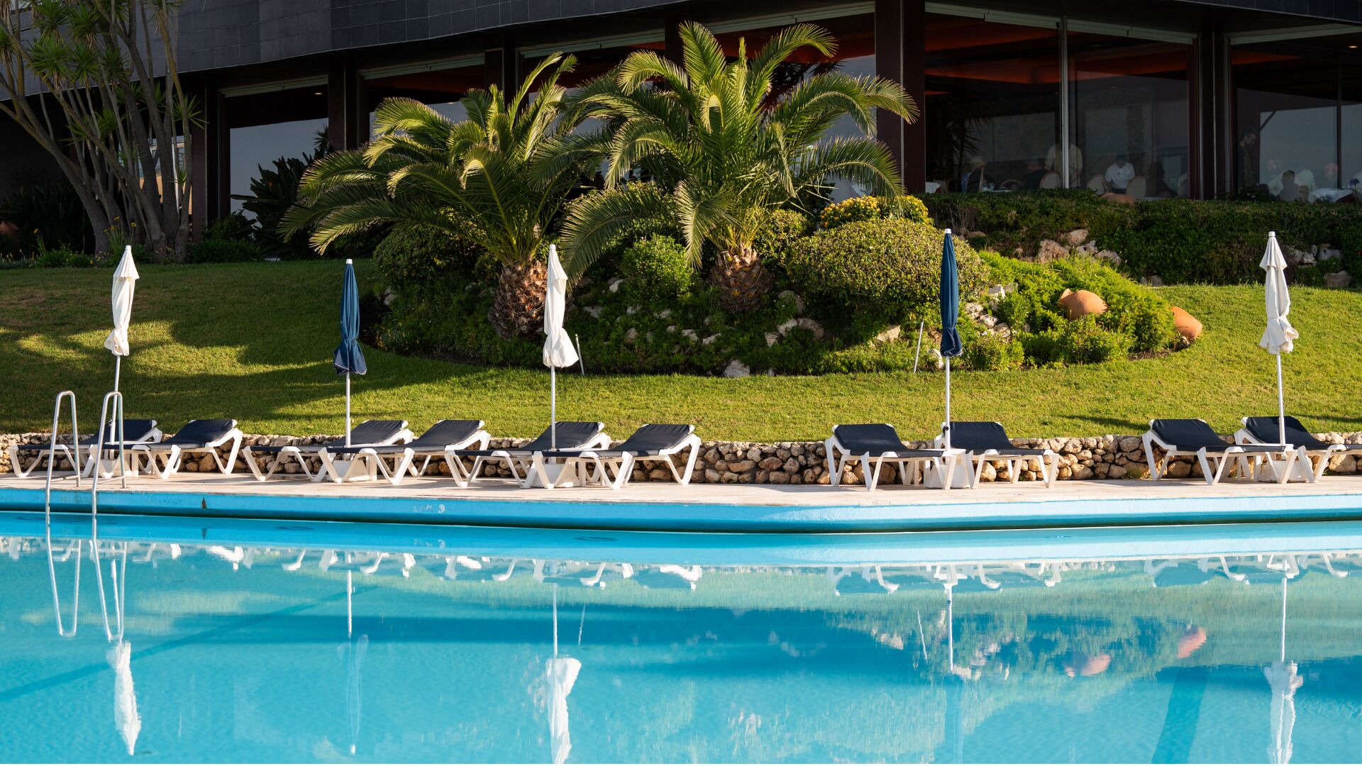 Área de piscina com espreguiçadeiras e guarda-sóis, cercada por vegetação e um edifício ao fundo.