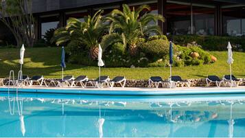 Área de piscina com espreguiçadeiras e guarda-sóis, cercada por vegetação e um edifício ao fundo.