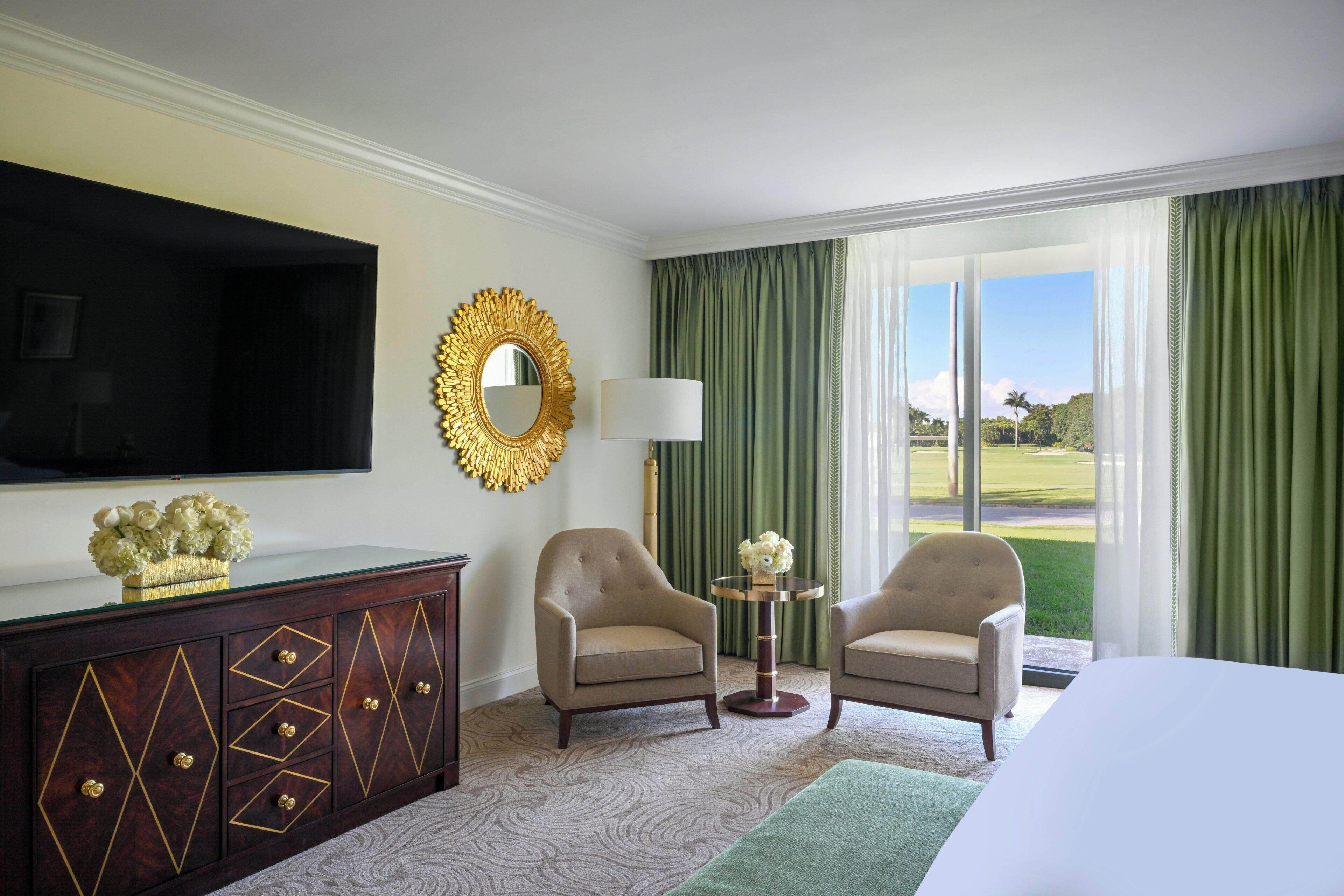 Quarto de hotel com uma televisão de tela plana grande, duas poltronas, uma mesinha com um vaso de flores, uma cama e vista para um campo de golfe através de grandes janelas.