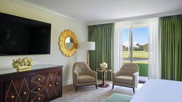 Quarto de hotel com uma televisão de tela plana grande, duas poltronas, uma mesinha com um vaso de flores, uma cama e vista para um campo de golfe através de grandes janelas.