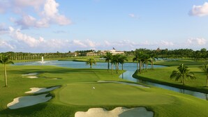 Um campo de golfe com bunkers de areia, lagoa e palmeiras.