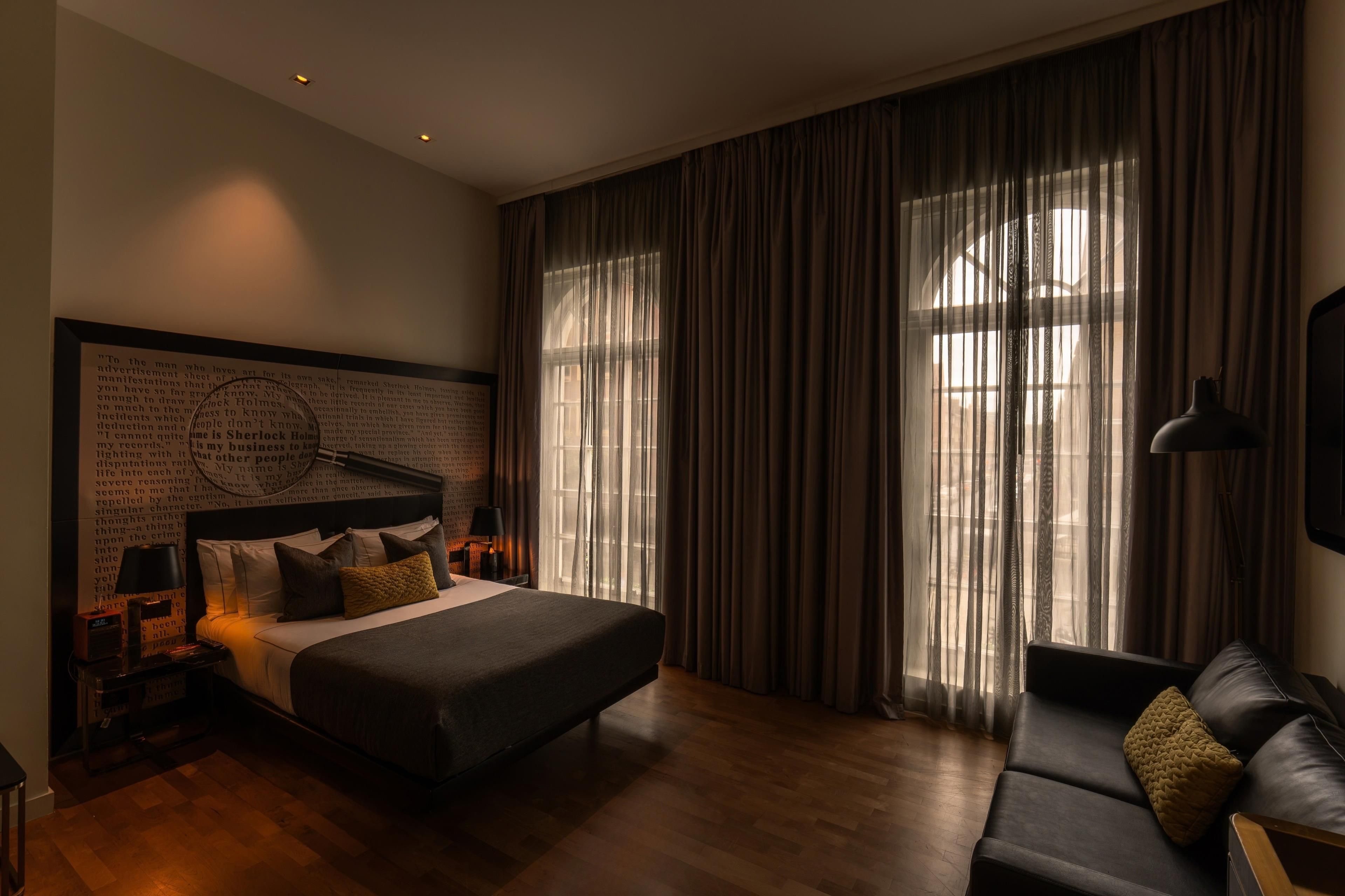 Quarto de hotel moderno com uma cama grande, um sofá e vista através de cortinas leves.