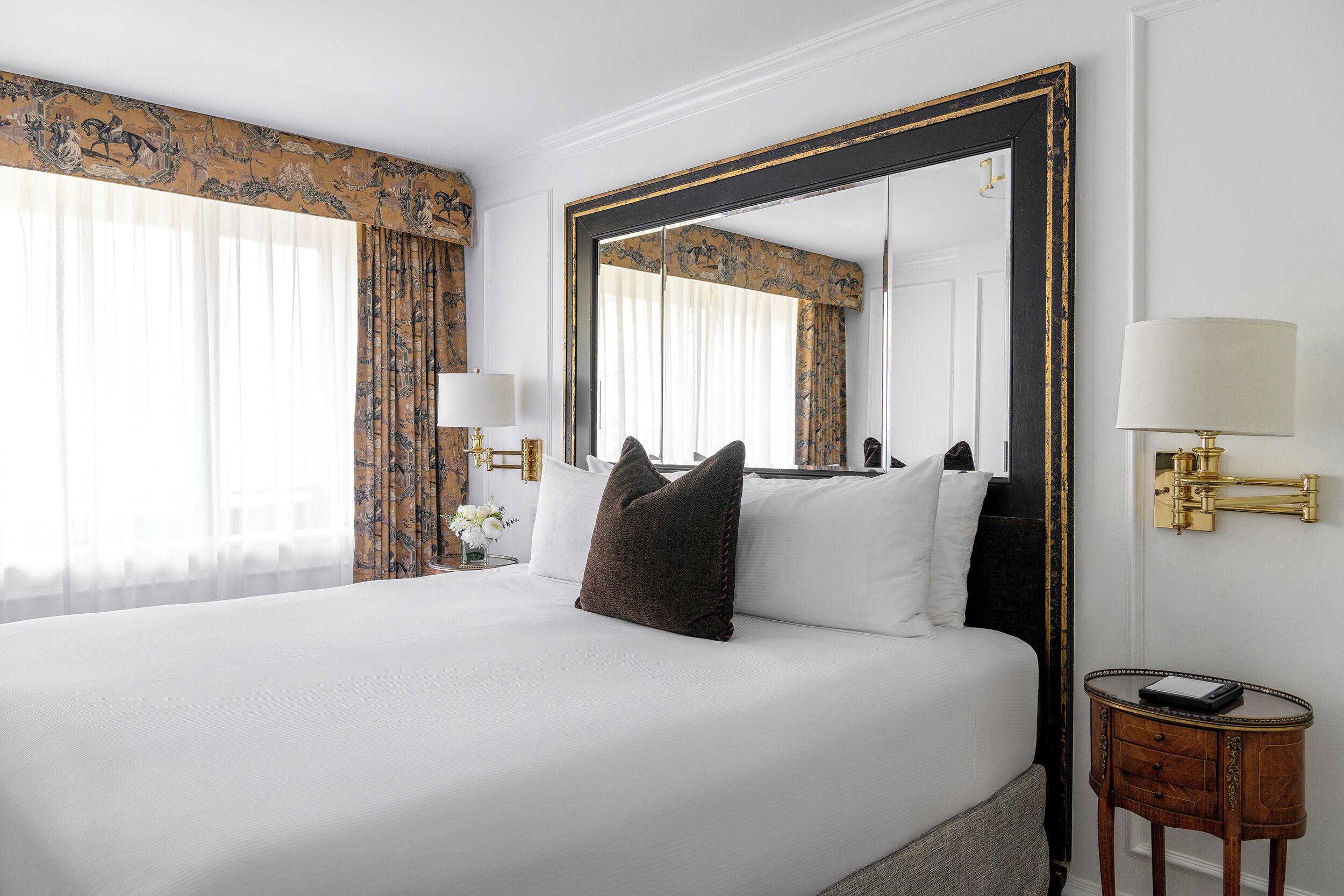 Quarto de hotel com uma cama grande, um espelho de corpo inteiro e uma pequena mesa de cabeceira com uma luminária.
