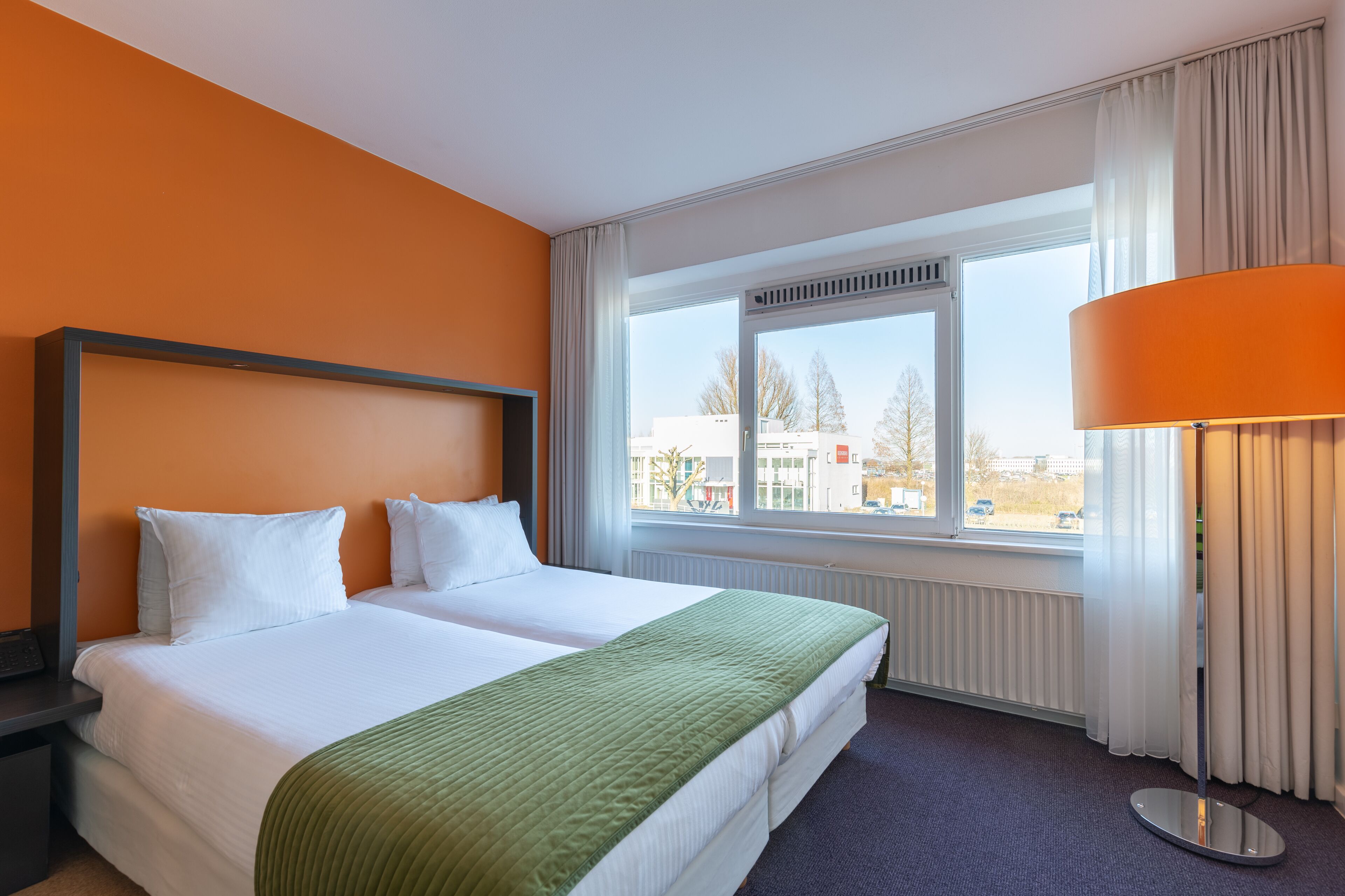 Quarto de hotel com duas camas, uma janela grande com cortinas, um abajur e vista para prédios.