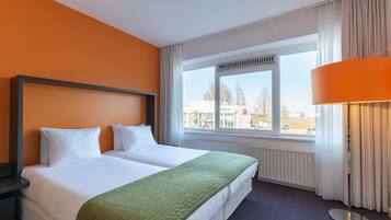 Quarto de hotel com duas camas, uma janela grande com cortinas, um abajur e vista para prédios.