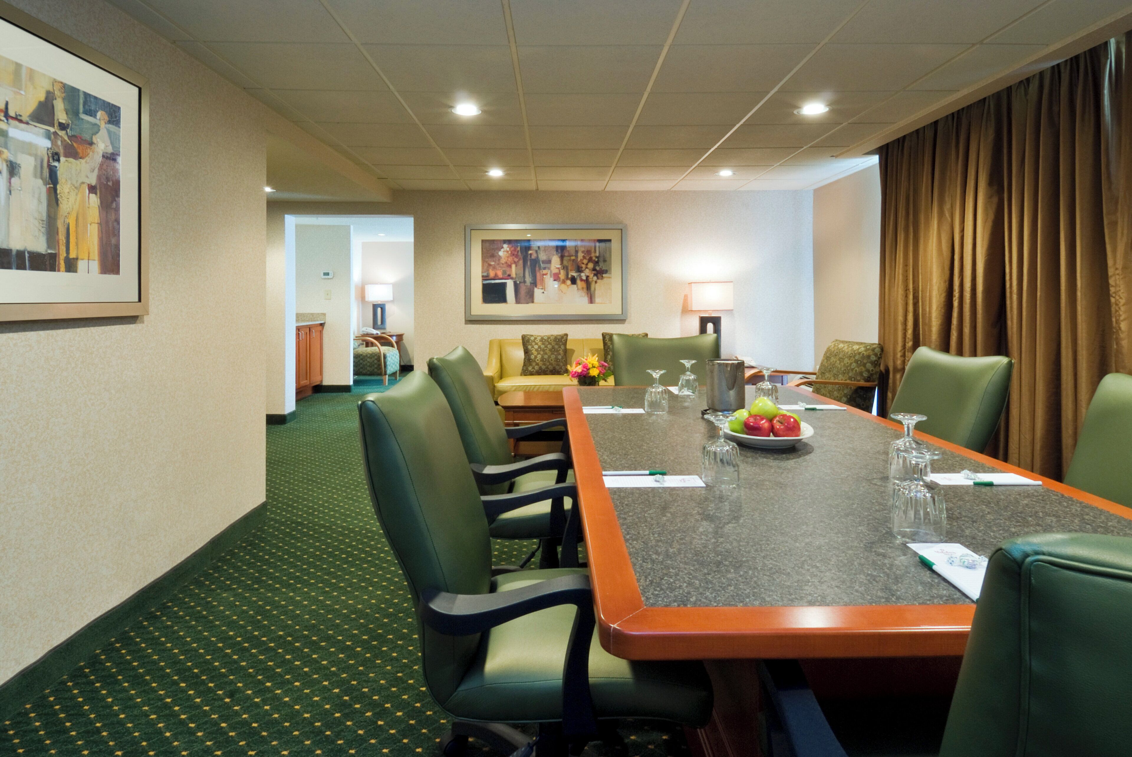 Sala de conferência com uma mesa comprida, cadeiras verdes, um quadro na parede e vista para um quarto de hotel.