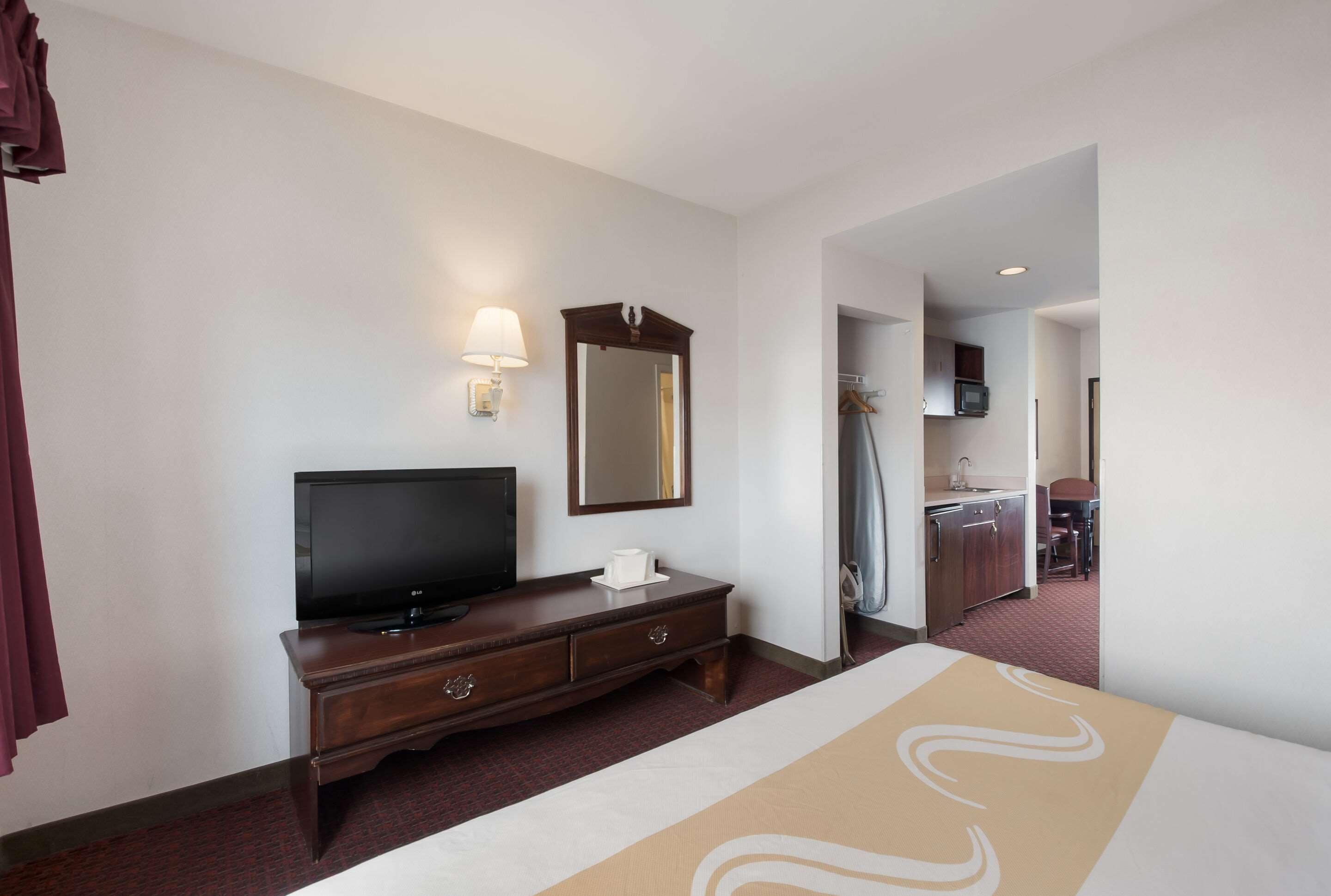Quarto de hotel com uma cama, uma televisão sobre uma cômoda de madeira, um espelho e uma cozinha compacta ao fundo.