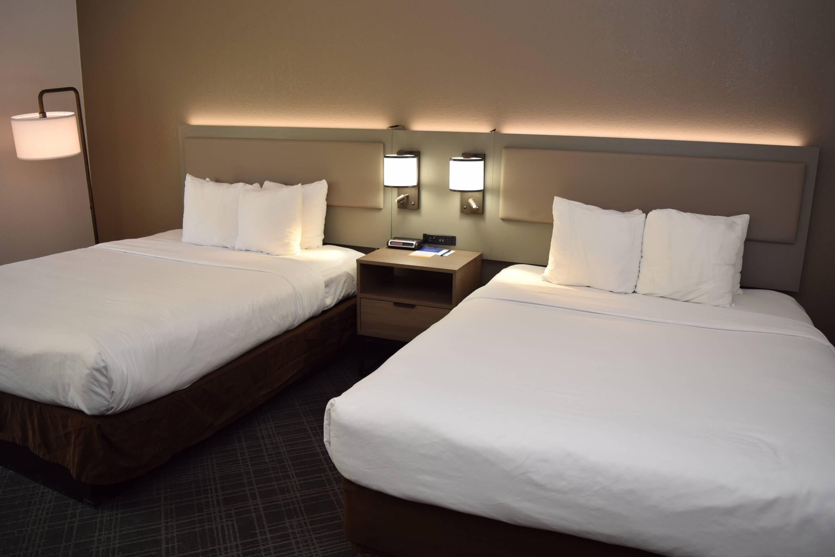 Quarto de hotel com duas camas de solteiro, mesa de cabeceira com luminária e luminária fixada na parede.