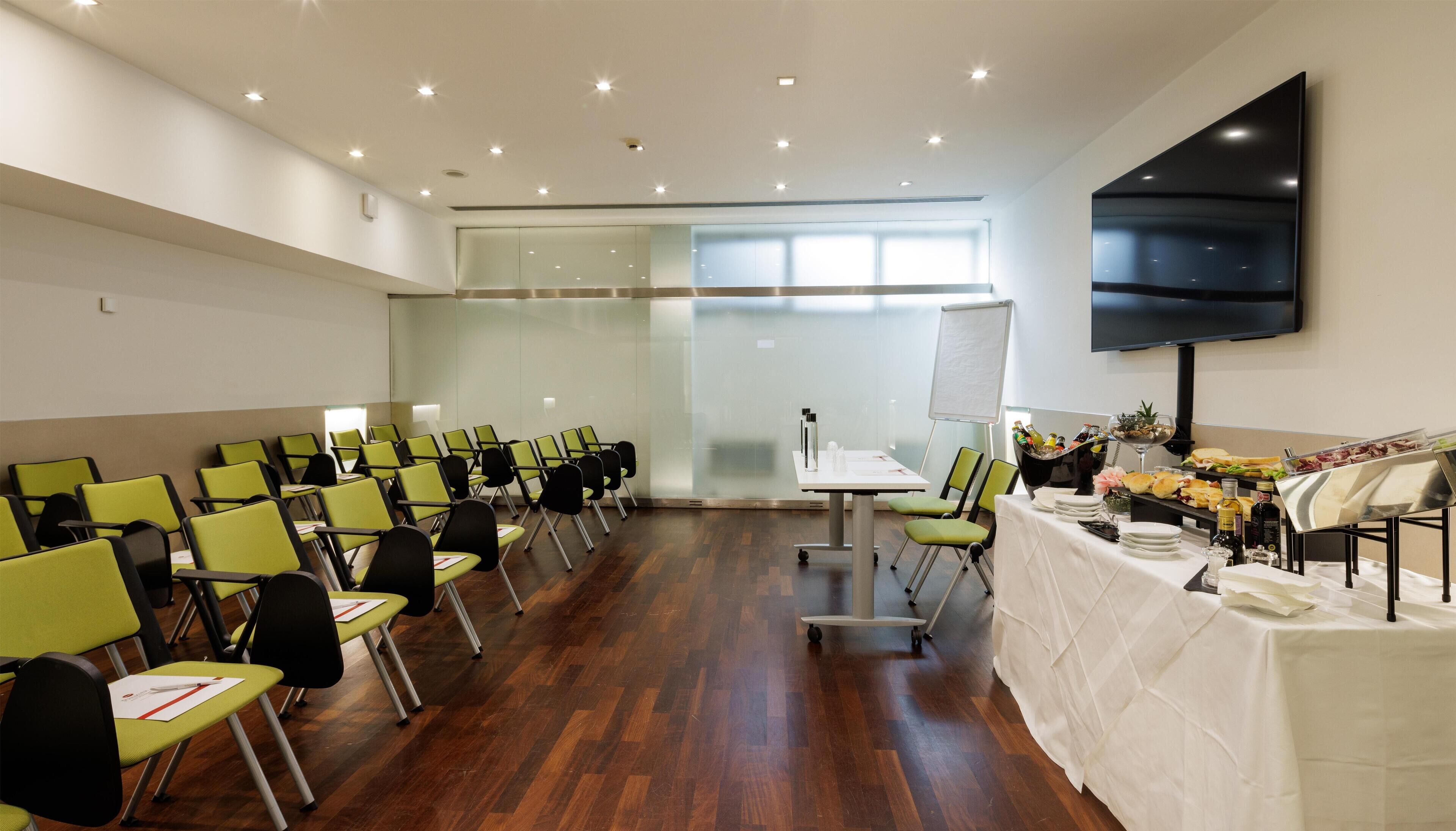 Sala de conferência com fileiras de cadeiras, um quadro branco, uma mesa com comida e bebida, e uma televisão na parede.