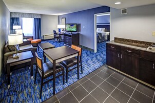 Quarto de hotel com área de refeições, televisão, sofá e cozinha compacta.