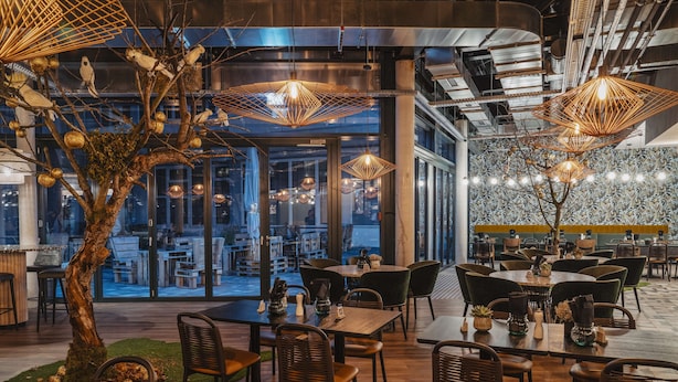 Interior moderno de restaurante com mesas de madeira, cadeiras de vime e luminárias pendentes decorativas.