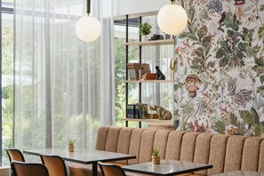 Uma área de estar aconchegante com sofá e mesas, uma estante com livros e objetos decorativos, e uma janela grande com cortinas leves.