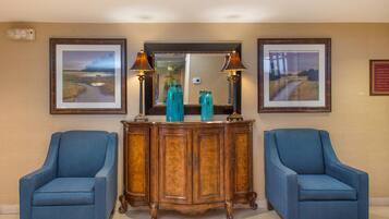 Um hall com duas poltronas azuis, uma mesa consola de madeira com objetos decorativos, quadros de paisagens emolduradas e um espelho sobre a mesa.