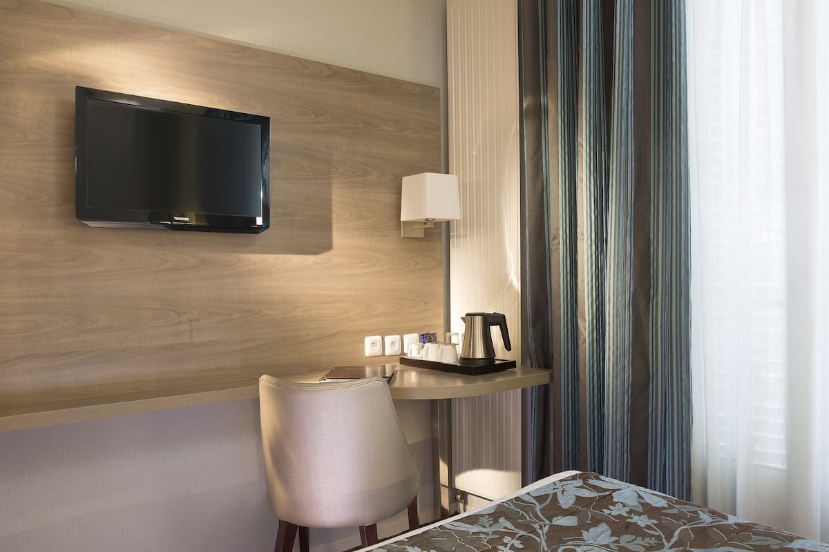 Quarto de hotel moderno com televisão de tela plana na parede, uma pequena escrivaninha com luminária, uma cadeira e uma cama com colcha estampada.