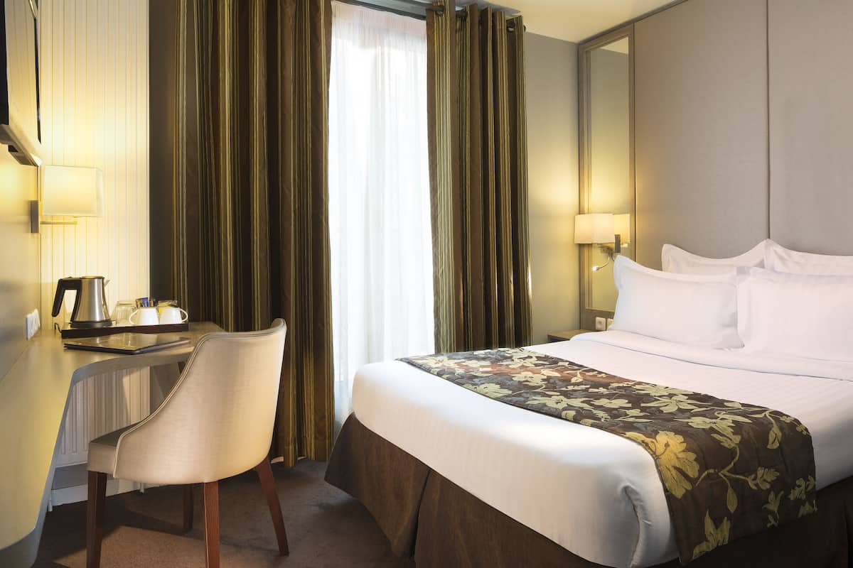 Quarto de hotel com cama, escrivaninha, cadeira e janela com cortinas.