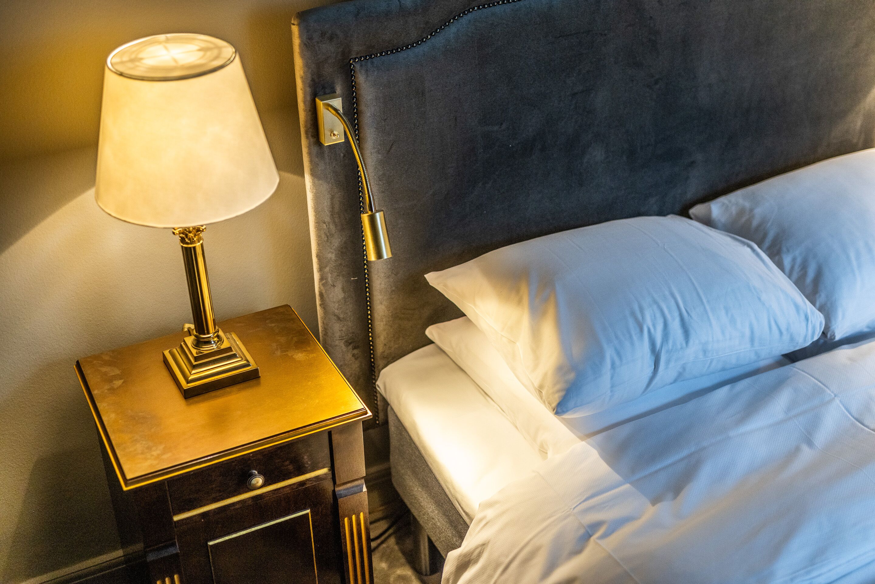 Uma cama bem arrumada com lençóis brancos e cabeceira, uma luminária de mesa com base dourada e uma pequena mesa de cabeceira de madeira com gaveta.