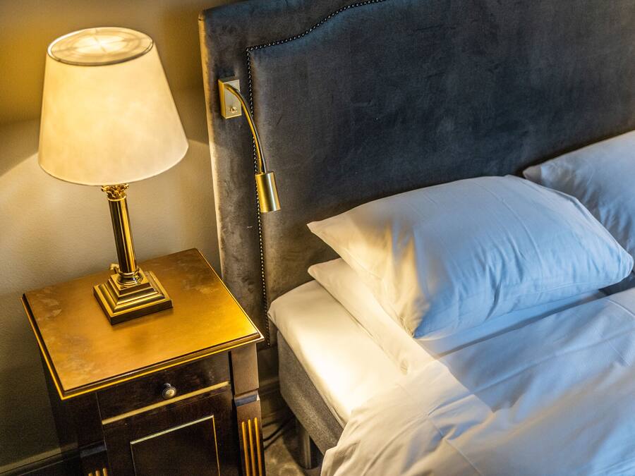 Uma cama bem arrumada com lençóis brancos e cabeceira, uma luminária de mesa com base dourada e uma pequena mesa de cabeceira de madeira com gaveta.