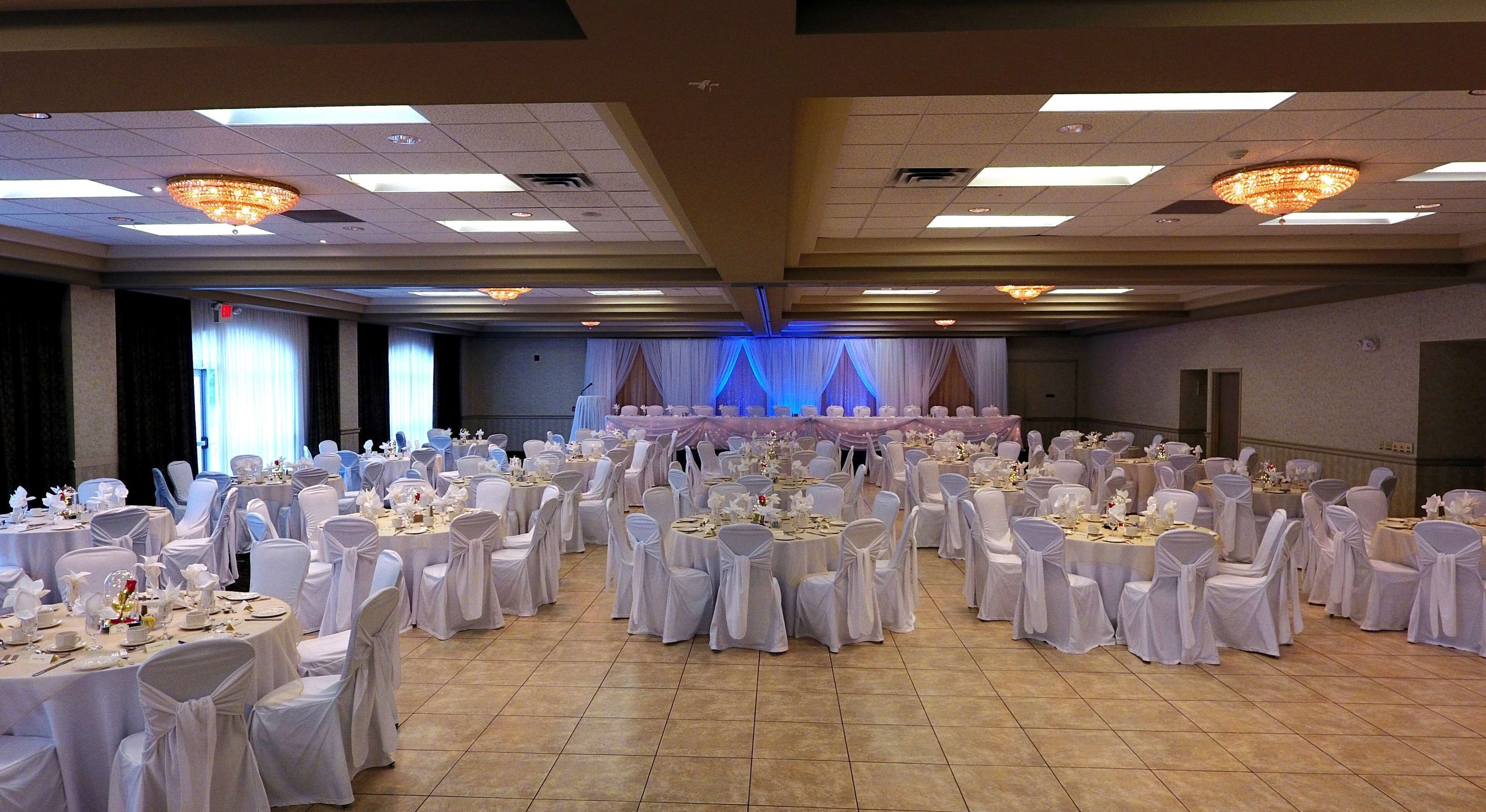 Um amplo salão de banquetes com mesas redondas cobertas por toalhas brancas e cadeiras, um palco com cortinas azuis e lustres no teto.