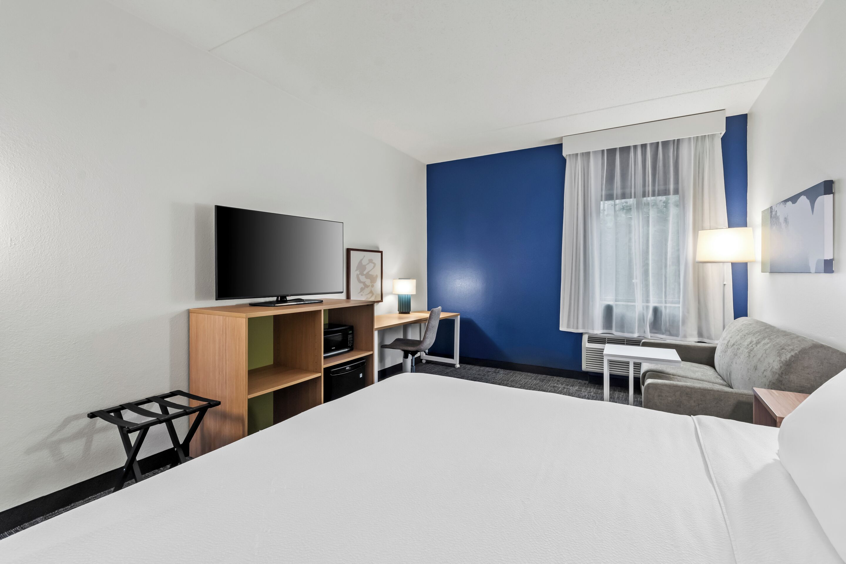 Quarto de hotel com uma cama grande, uma escrivaninha com cadeira, televisão, mesa pequena e um sofá.