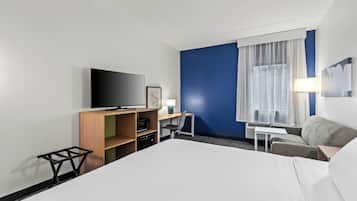 Quarto de hotel com uma cama grande, uma escrivaninha com cadeira, televisão, mesa pequena e um sofá.