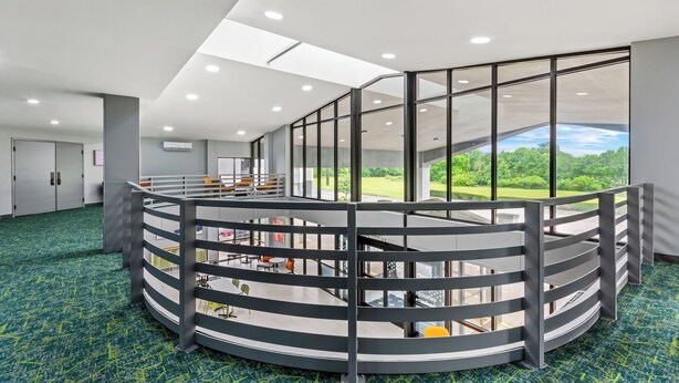 Um lobby de hotel moderno com guarda-corpo de vidro curvo, vista para o exterior e chão coberto por carpete verde.