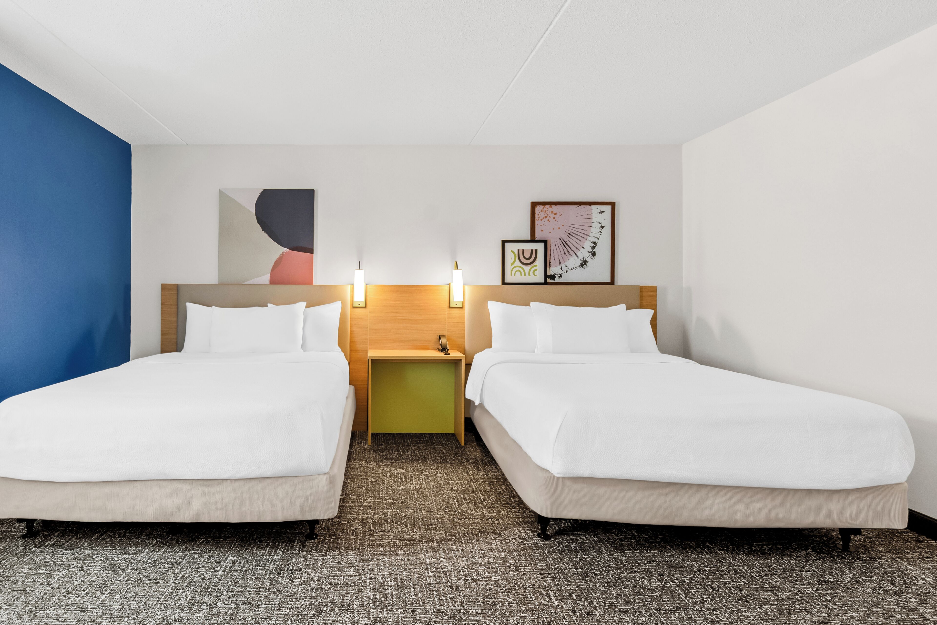 Quarto de hotel com duas camas, uma pequena mesa de cabeceira e luminária fixada na parede.