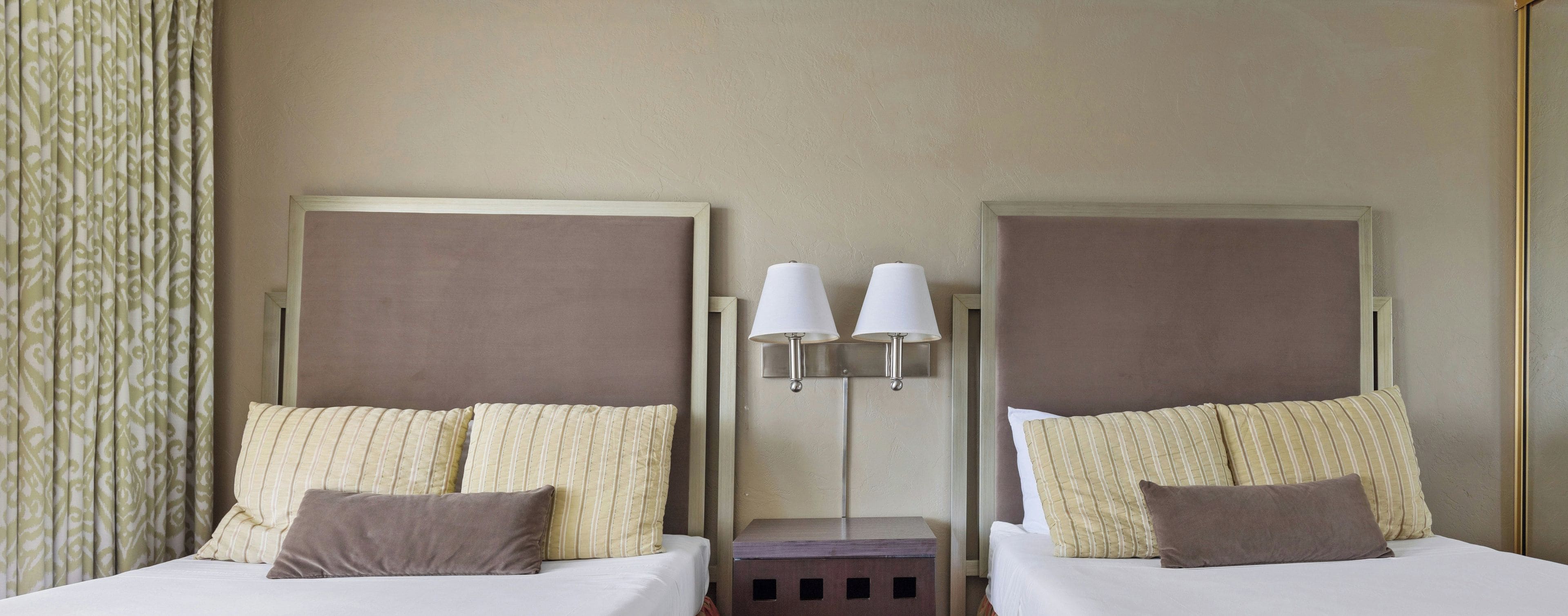 Quarto de hotel com duas camas, cada uma com cabeceira, mesa de cabeceira e duas luminárias fixadas na parede.