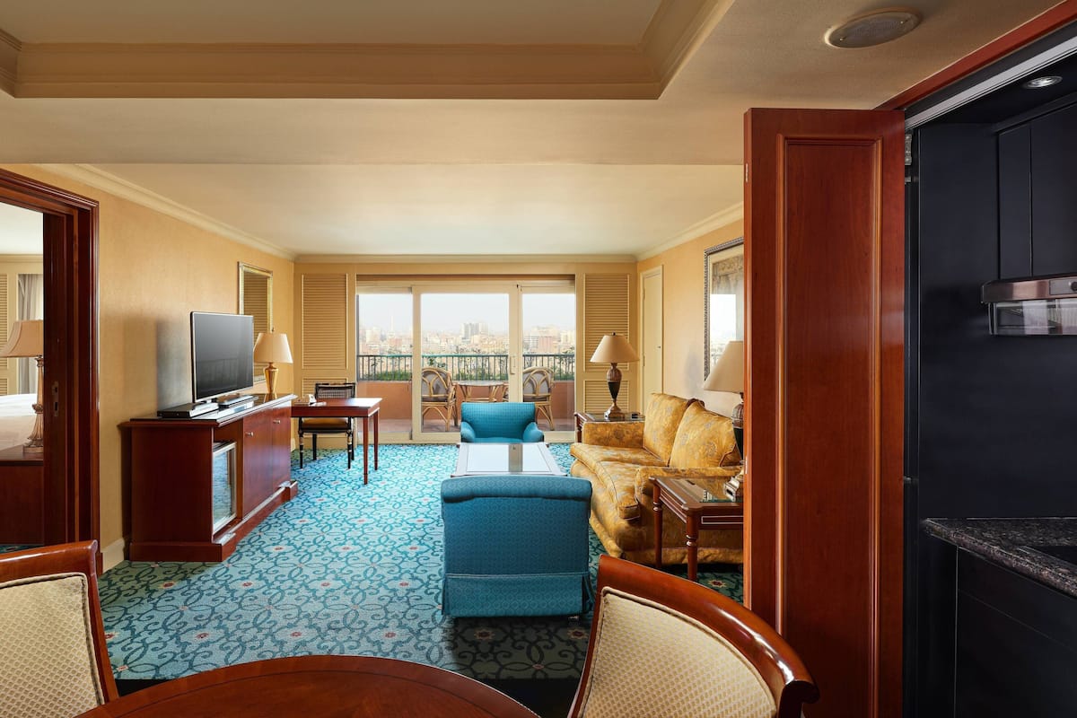Quarto de hotel com uma grande janela, televisão, sofá, mesa de centro e área de refeições.