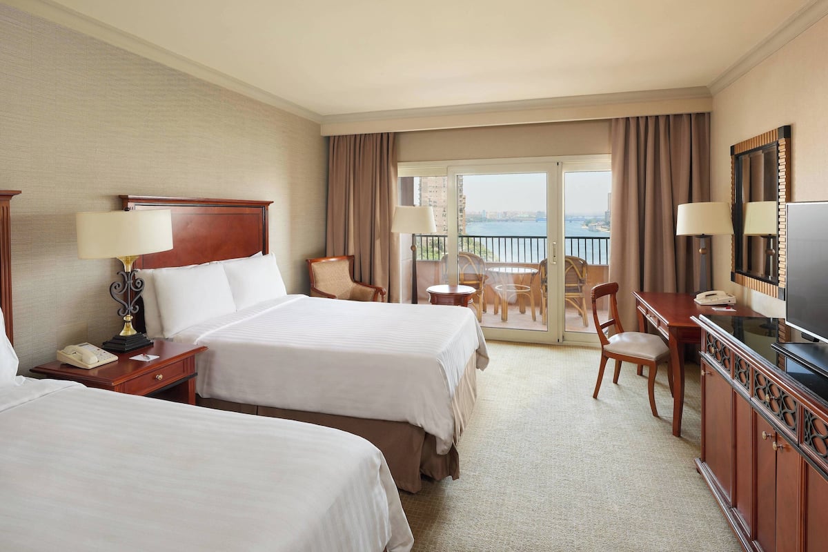 Quarto de hotel com duas camas, uma mesa com uma TV, uma cadeira e vista da cidade pelas portas da varanda.
