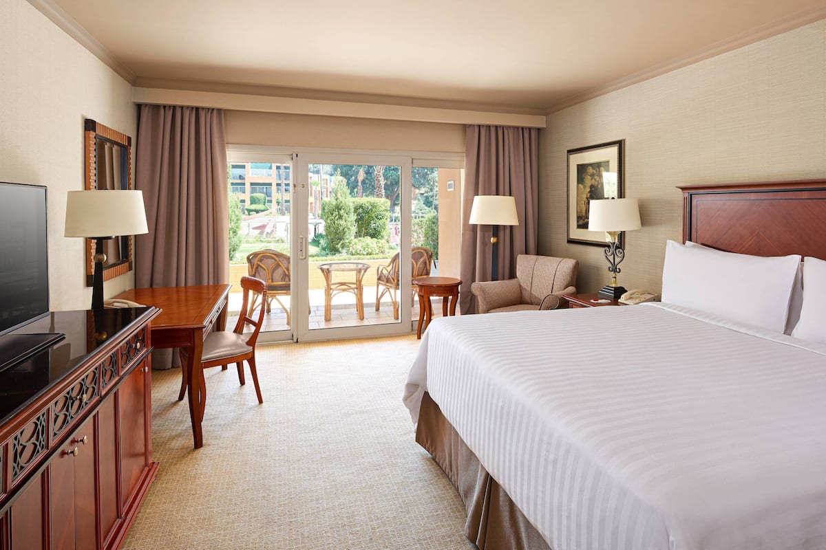 Quarto de hotel com uma cama grande, uma mesa com TV, uma cadeira, uma mesinha e vista para o exterior por uma porta de correr.