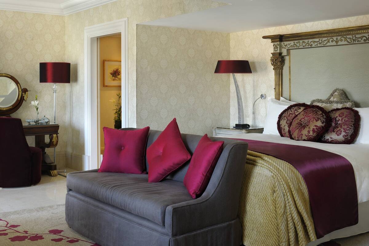 Quarto de hotel com sofá cinzento, almofadas vermelhas, uma cama grande com colcha dourada e vermelha, um espelho grande e duas luminárias.