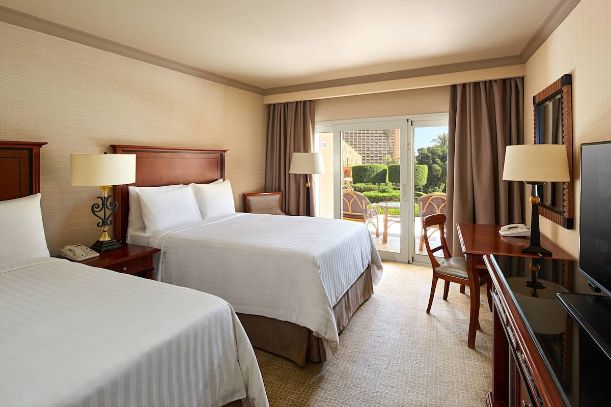 Quarto de hotel com duas camas, uma escrivaninha com cadeira, televisão e vista para o exterior.