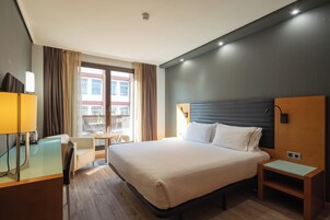 Quarto de hotel com uma cama grande, uma mesa com abajur, uma mesinha e vista para o exterior.
