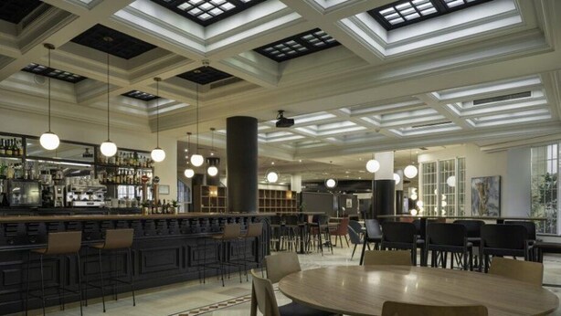 Um espaço amplo de bar, com teto alto e claraboias, rodeado por assentos e um balcão com bancos.