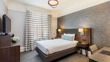 Quarto de hotel com uma cama grande, uma escrivaninha, uma cadeira, uma televisão e um abajur.