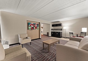 Quarto de hotel com televisão de tela plana na parede, um sofá, uma poltrona, uma mesinha e uma área de cozinha compacta.