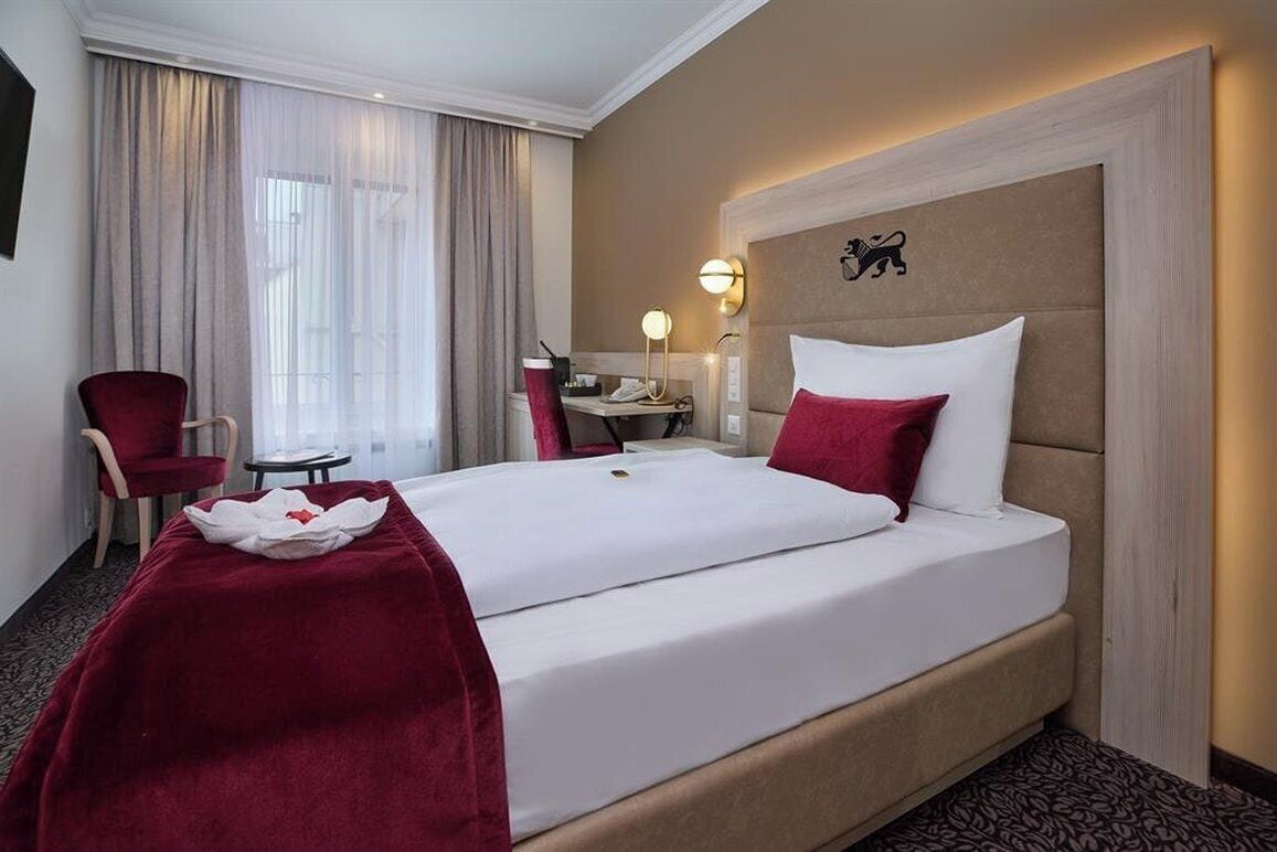 Quarto de hotel com cama, escrivaninha, cadeira e uma janela grande com cortinas.