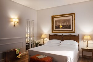 Quarto de hotel com uma cama grande, duas luminárias de cabeceira, um quadro histórico emoldurado, uma porta com painéis de vidro e uma mala no chão.