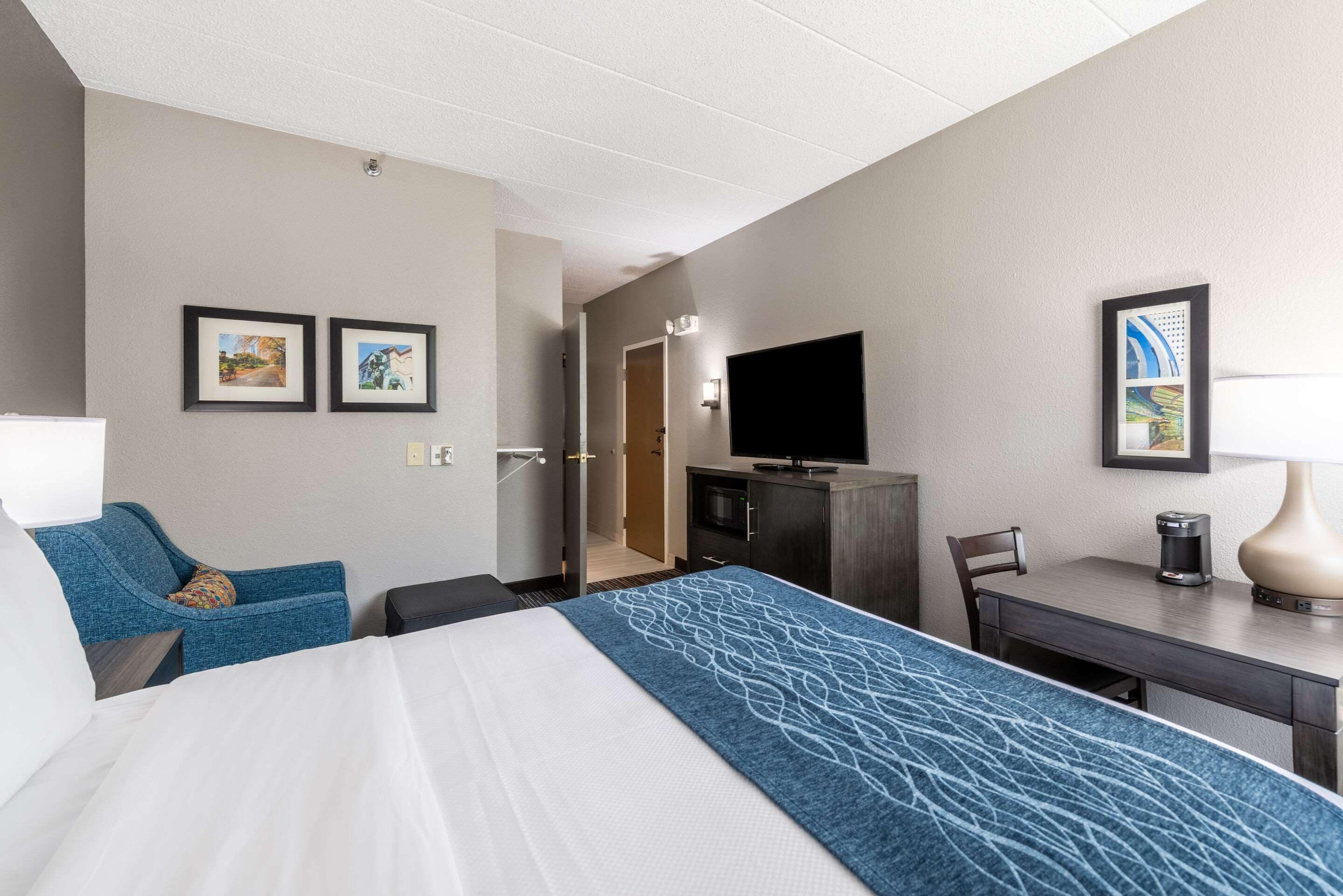Quarto de hotel com cama, escrivaninha, cadeira, televisão e quadros na parede.