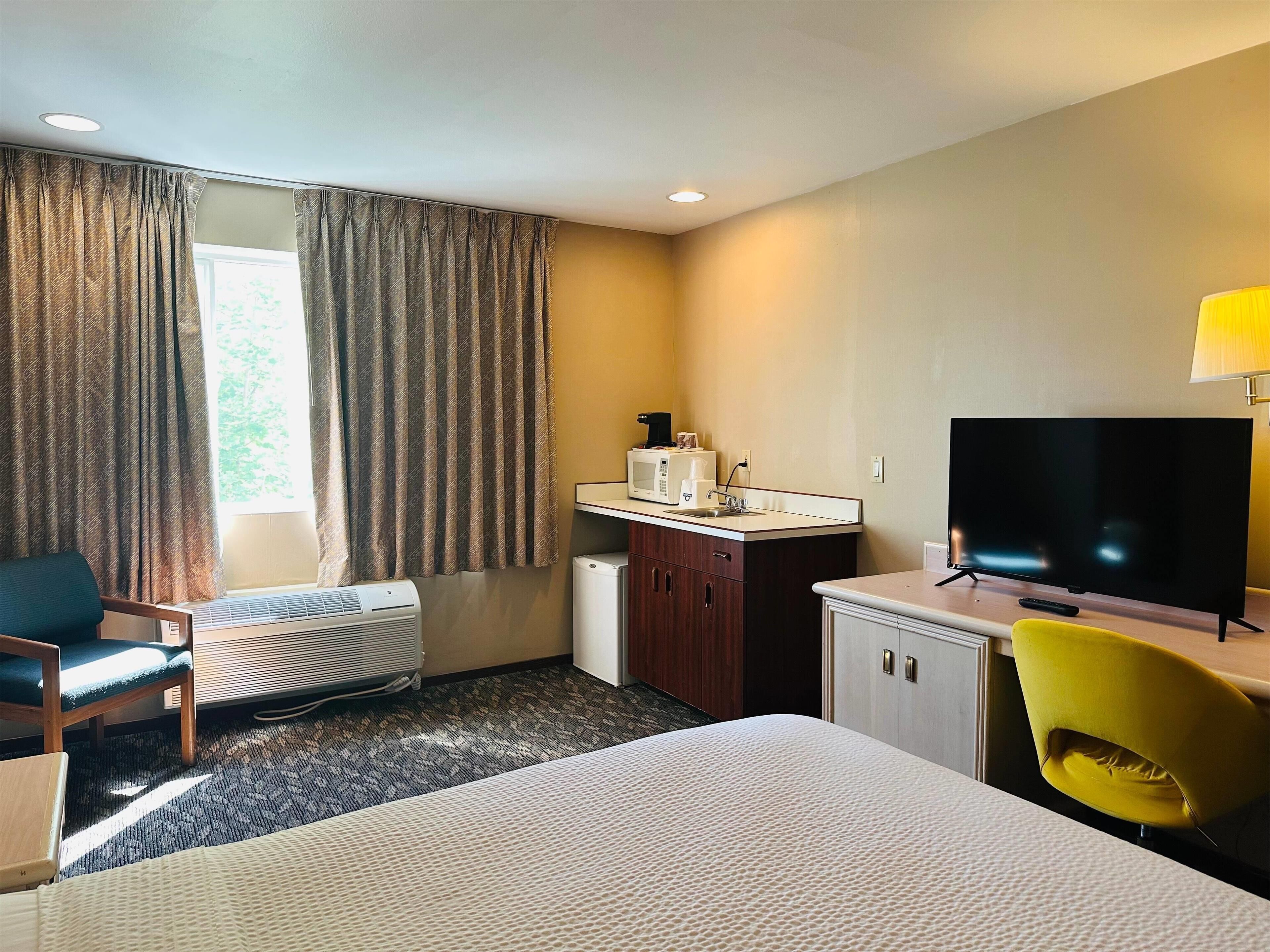 Quarto de hotel com cama, escrivaninha com televisão, cadeira, pequeno espaço de cozinha e janela com cortinas.