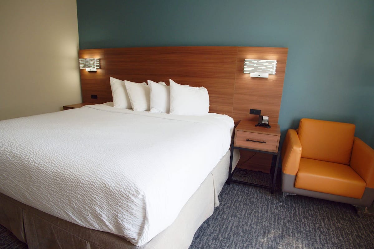Quarto de hotel com cama, mesa de cabeceira, poltrona e cabeceira com luzes fixadas na parede.