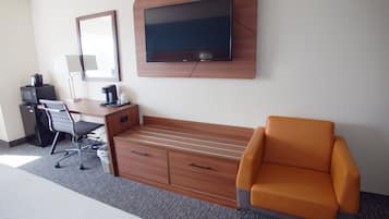 Quarto de hotel com cama, escrivaninha com computador, cafeteira, televisão na parede e uma poltrona laranja.