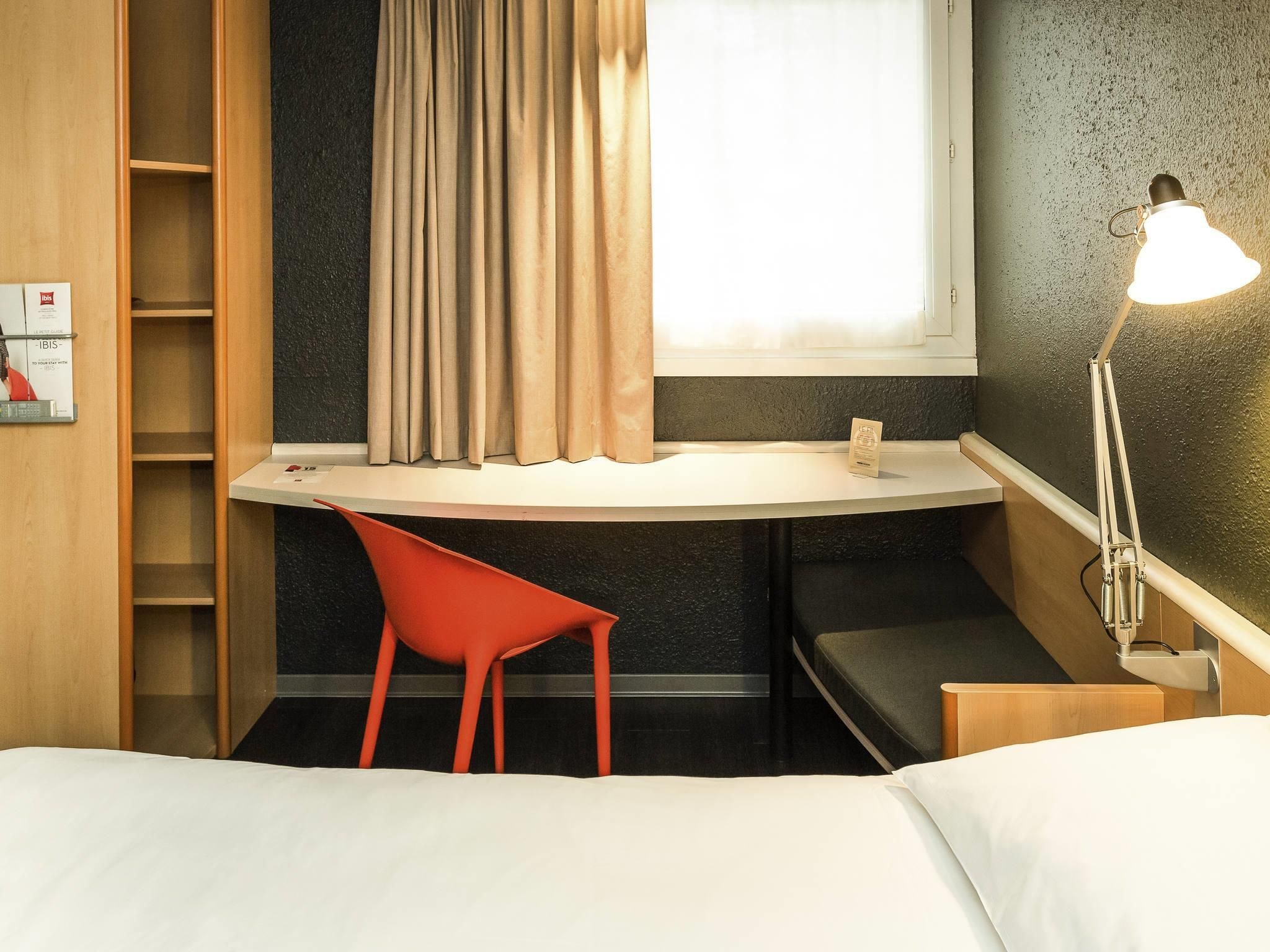 Quarto de hotel com escrivaninha, uma cadeira vermelha, cama, janela com cortinas e uma luminária.